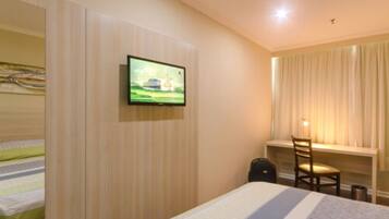 Quarto de hotel com cama, escrivaninha, cadeira, televisão e janela com cortinas.