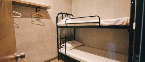 Quarto pequeno com uma cama de solteiro, piso de madeira, uma cama de metal e uma prateleira de madeira com cabides.