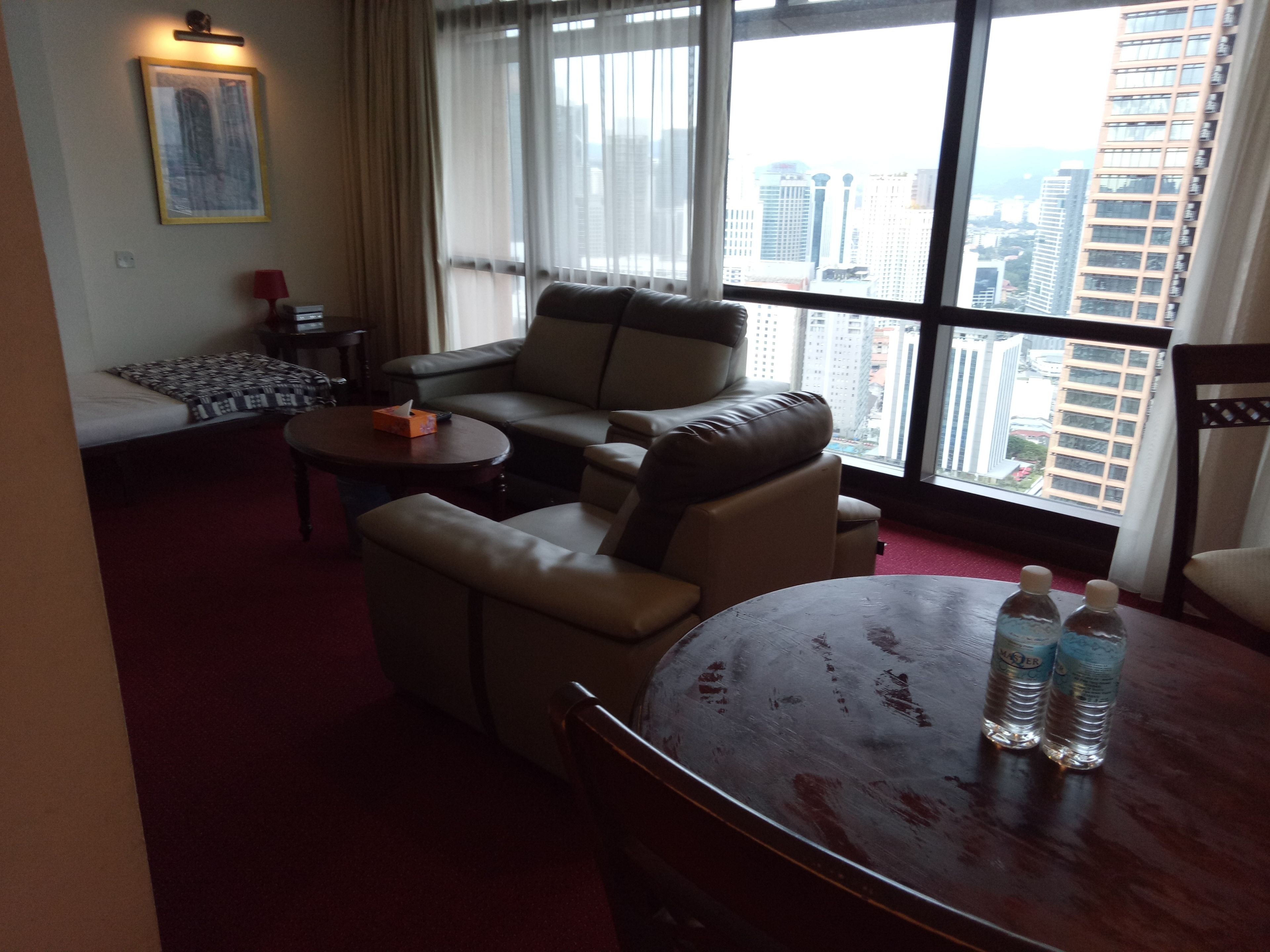 Quarto de hotel com sofá, poltronas, uma mesinha e vista da cidade através de grandes janelas.