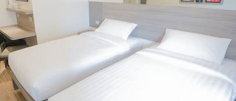 Quarto de hotel com duas camas de solteiro, mesa de cabeceira, cadeira, escrivaninha pequena, espelho e um quadro de avisos fixado na parede.