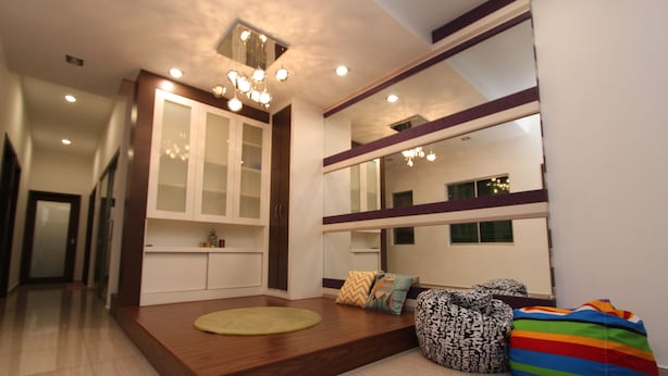 Uma sala de estar moderna com uma plataforma de madeira, um sofá, um pufe colorido e um espelho grande refletindo o ambiente.