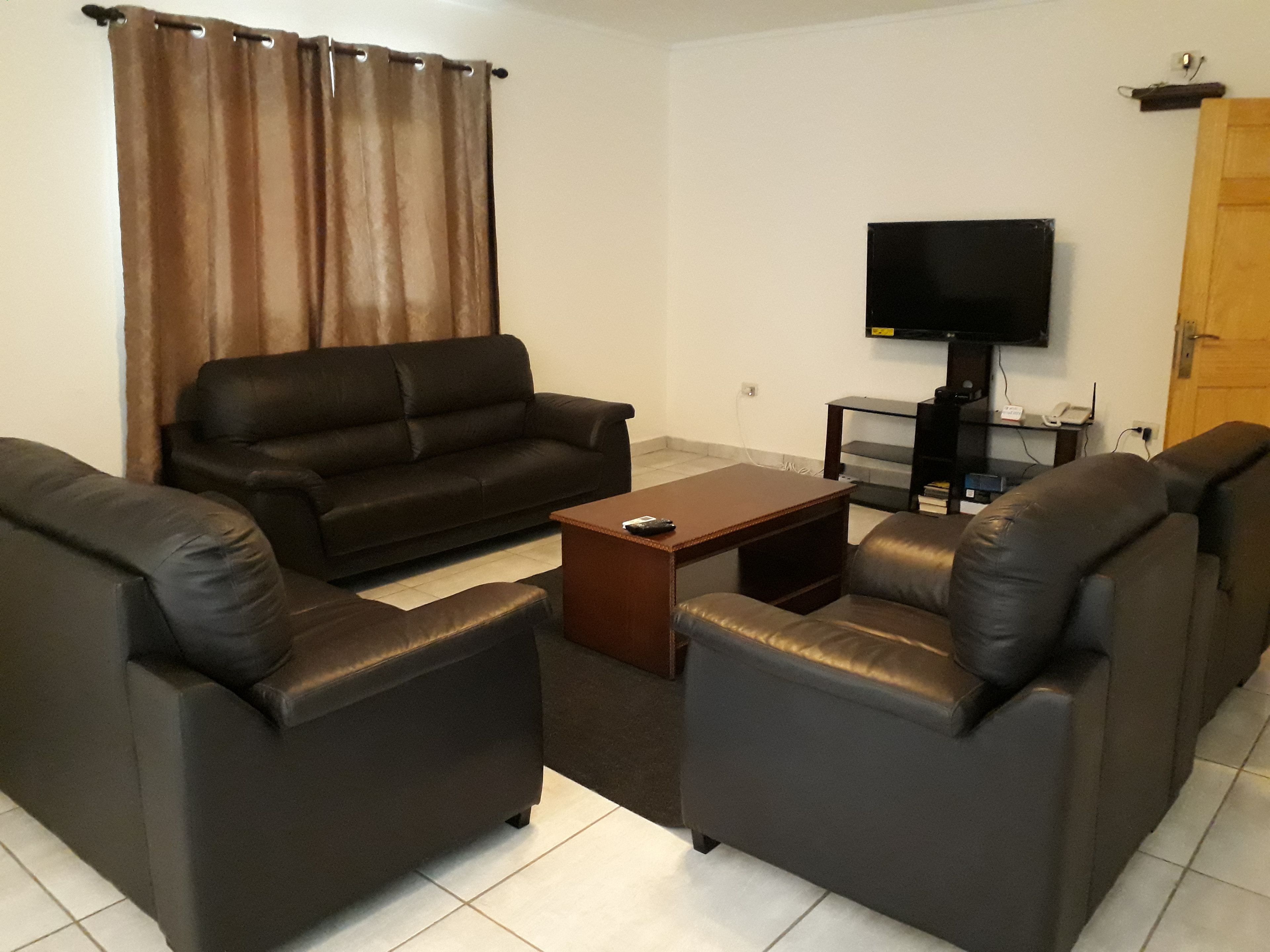 Uma sala de estar com um sofá preto, duas poltronas, uma mesa de centro e uma TV sobre um suporte.