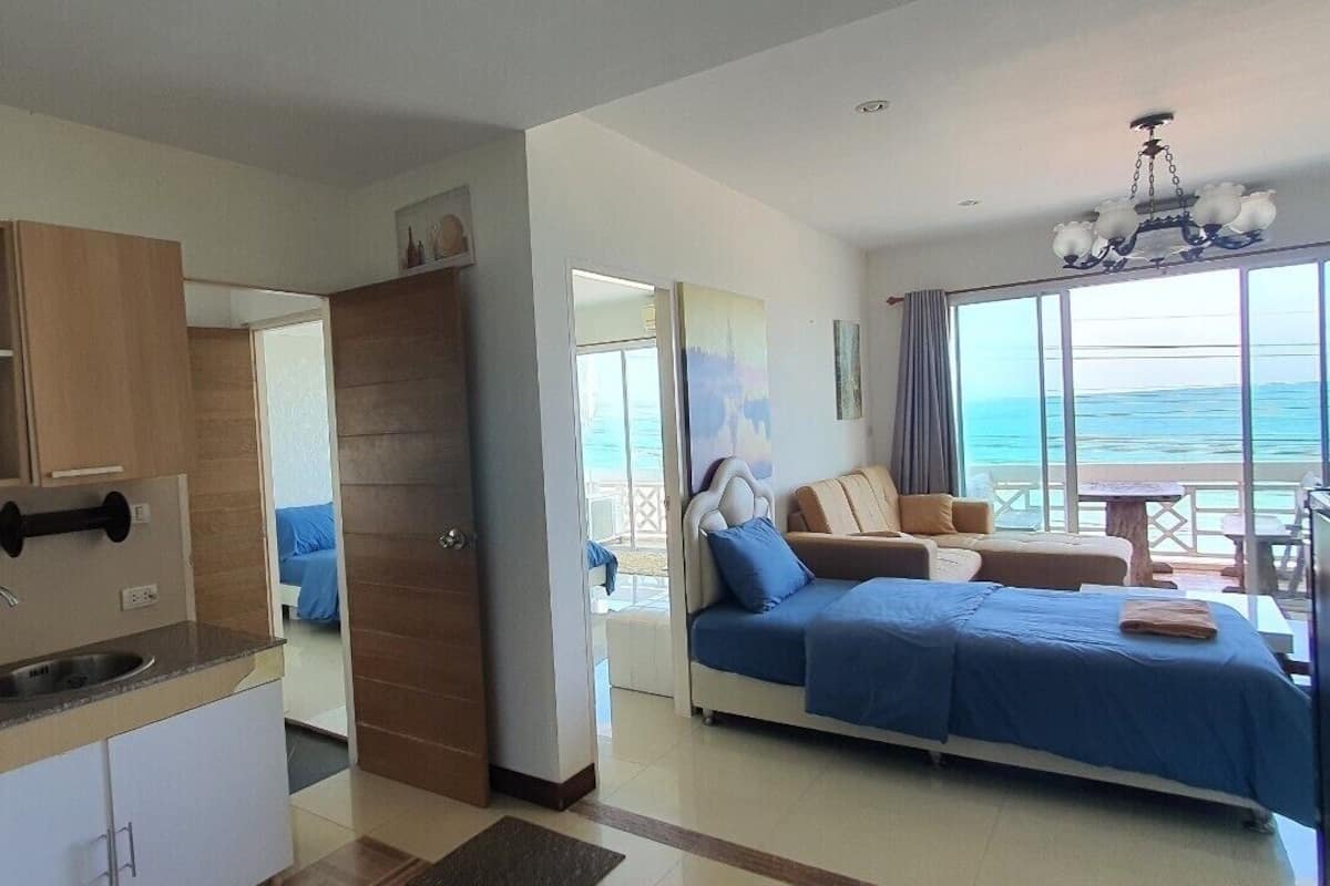 Quarto de hotel moderno com cozinha compacta, cama com roupa de cama azul, área de estar com vista para o mar e varanda com mesa e cadeiras.