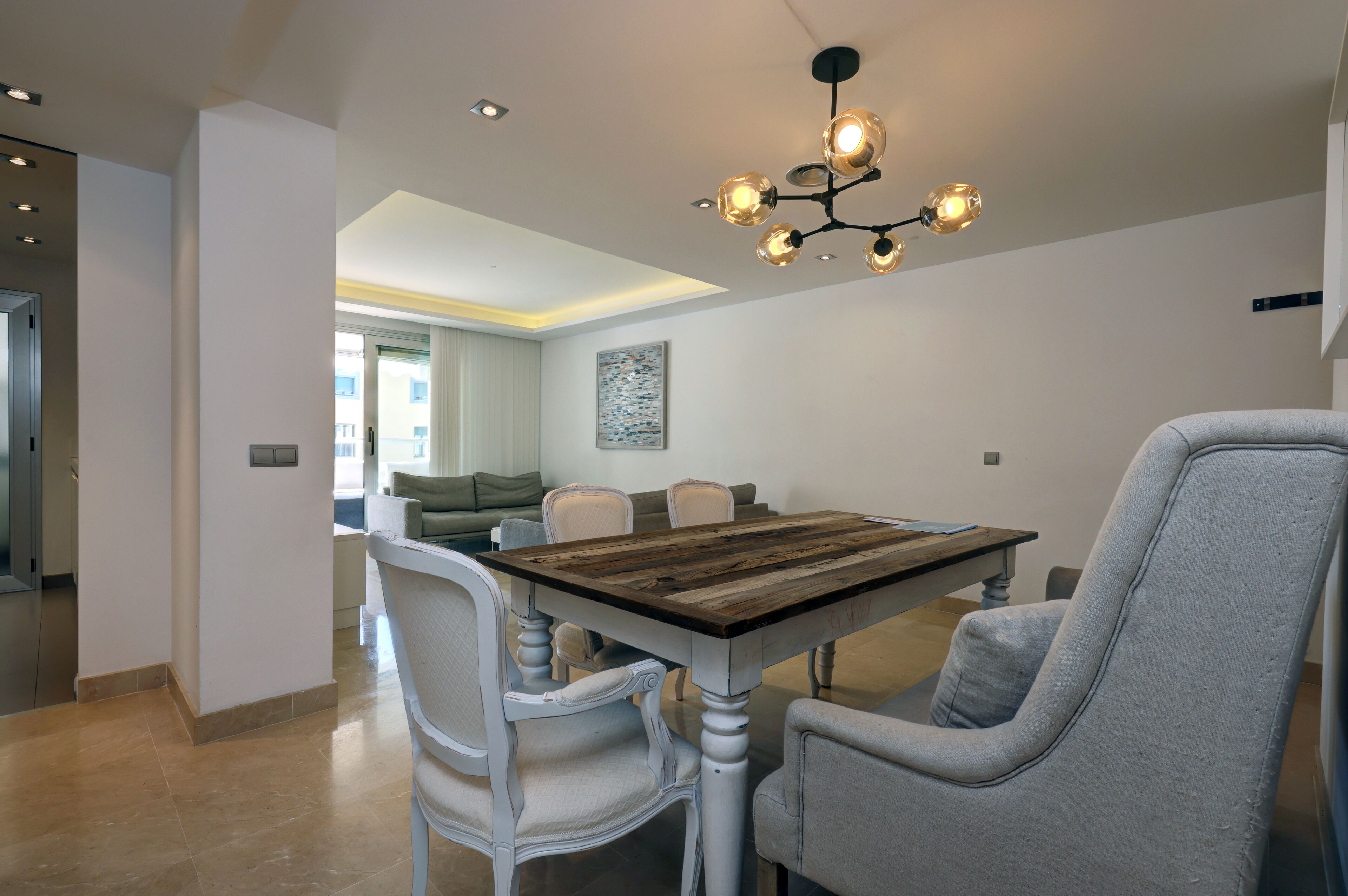 Uma sala de jantar com mesa de madeira e cadeiras estofadas, um lustre e vista para outro ambiente com sofá e pintura.