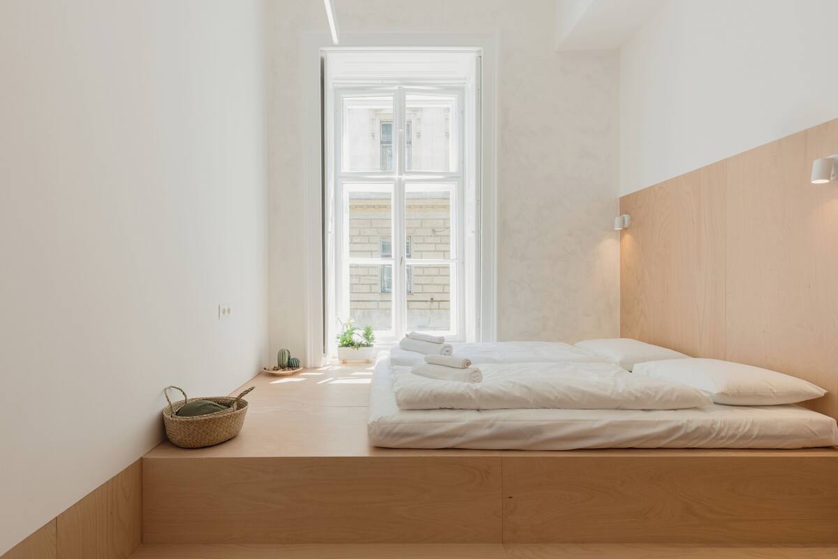 Quarto minimalista com uma cama em plataforma de madeira, uma cesta pequena de palha e um vaso com planta.
