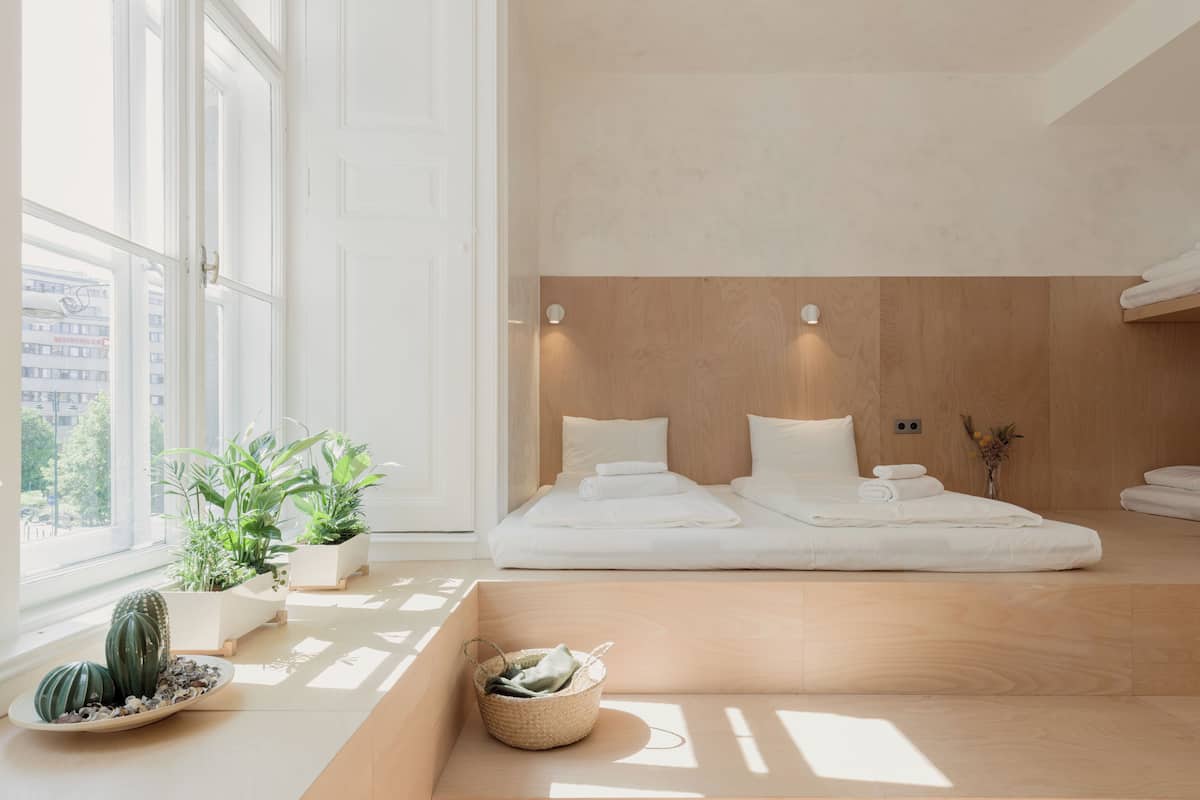Quarto minimalista com uma cama grande, cabeceira de madeira, plantas em vasos e vista da cidade através de grandes janelas.