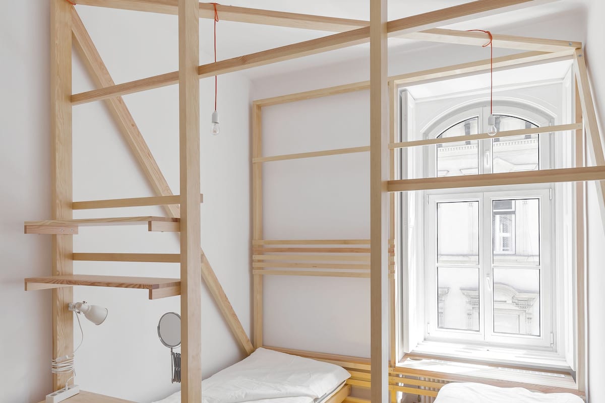 Um quarto com uma cama de solteiro, uma escrivaninha com cadeira, uma mesa pequena e uma janela com vista para edifícios.