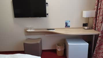 Quarto de hotel com cama, escrivaninha, televisão de tela plana, lixeira, mini-geladeira e luminária.