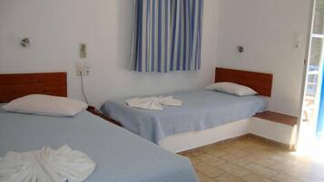 Quarto com duas camas, cada uma com cabeceira de madeira e colcha azul. Cortina listrada na janela, e uma toalha branca sobre a cama.
