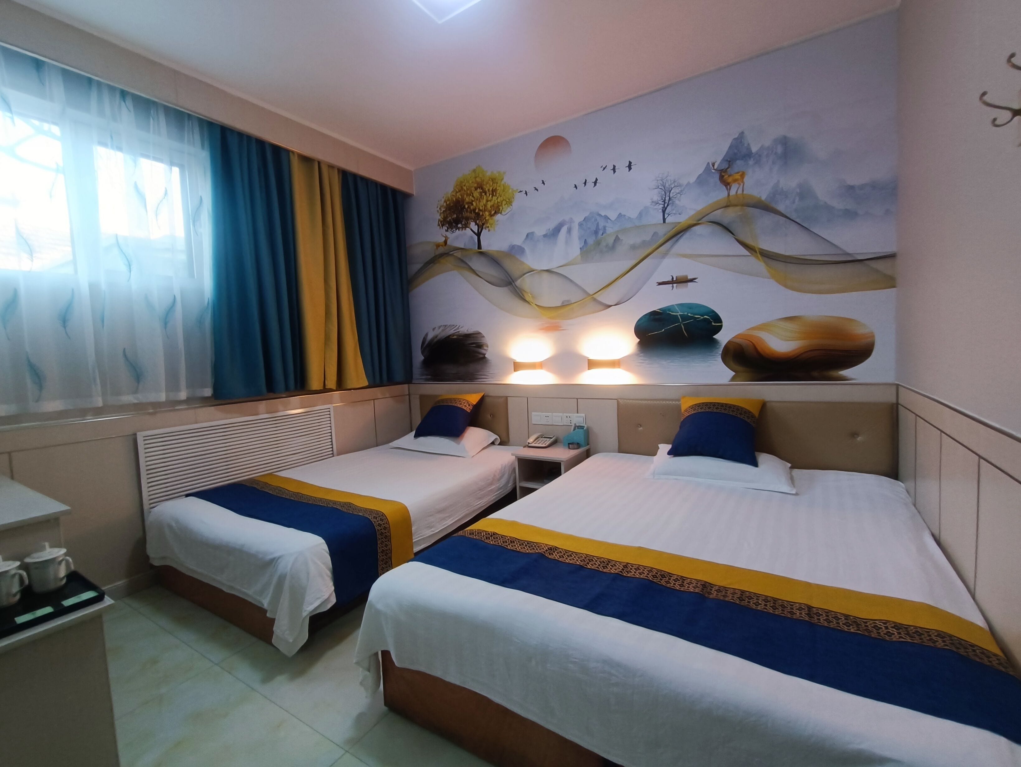 Quarto de hotel com duas camas, mesa de cabeceira, um mural na parede e janela com cortinas.
