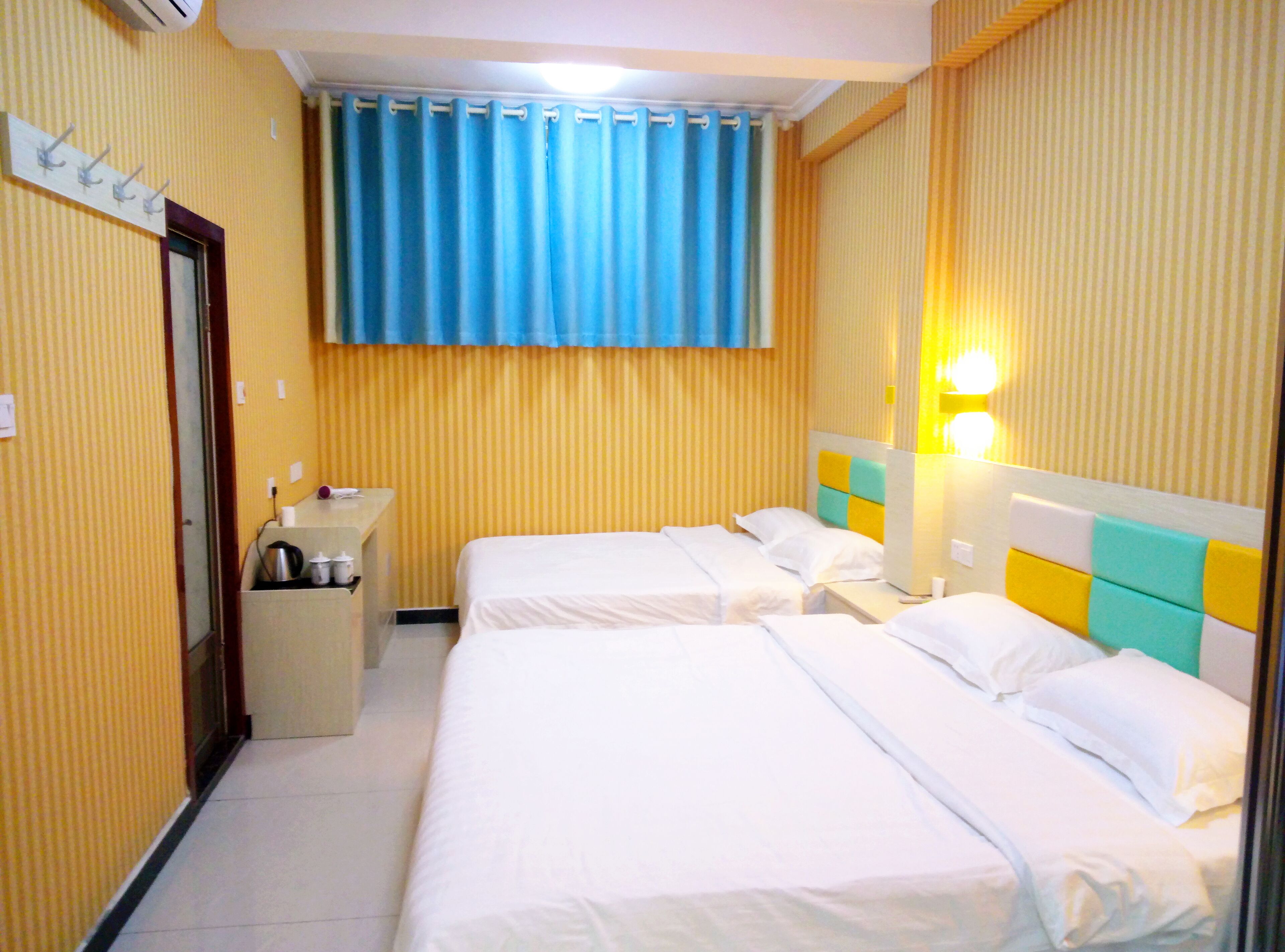Quarto de hotel com duas camas, uma pequena mesa de cabeceira, um criado-mudo, uma janela com cortinas azuis e um luminário fixado na parede.