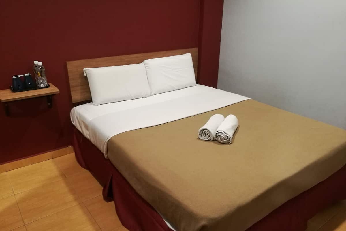 Uma cama bem arrumada com roupa de cama branca e castanha, uma cabeceira de madeira e uma pequena estante com uma garrafa de água e copos.