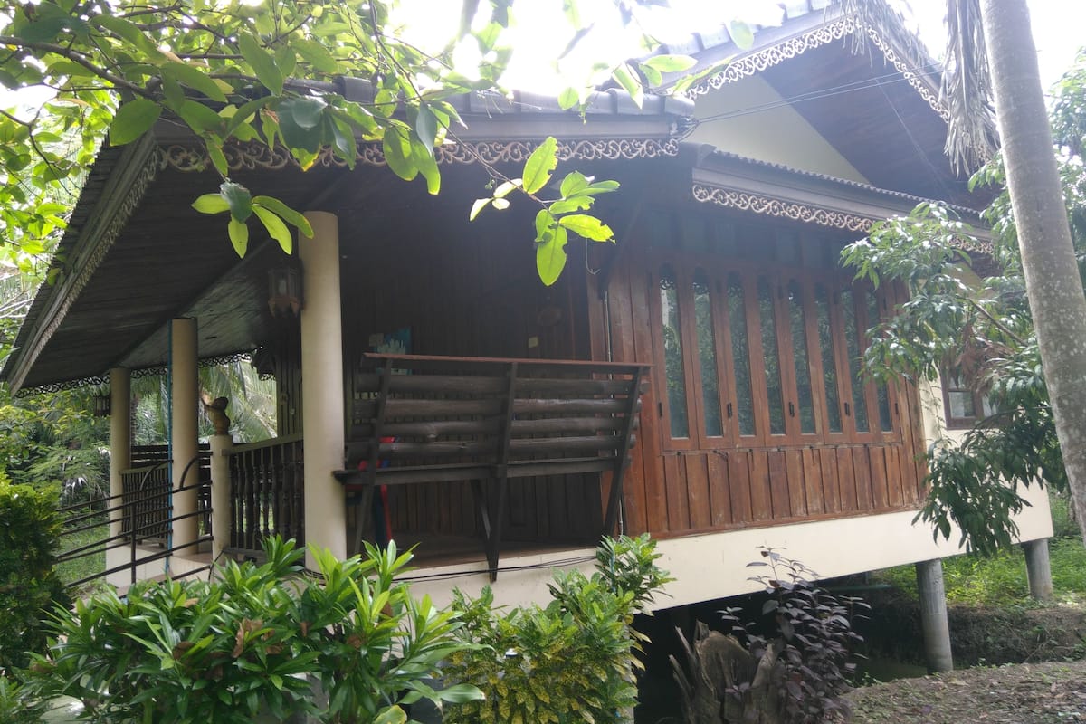 Uma casa de madeira tradicional com varanda, rodeada por vegetação exuberante.