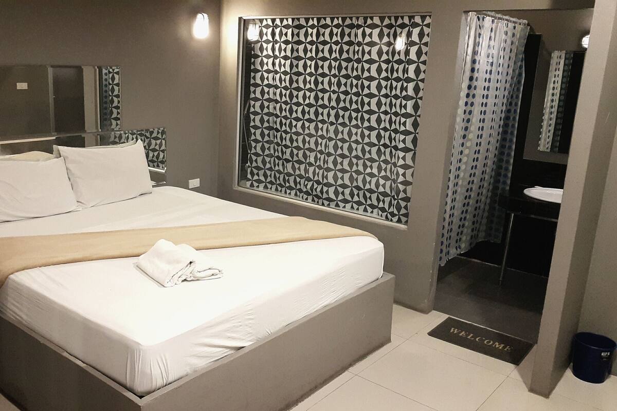 Quarto de hotel com uma cama grande, uma mesa de cabeceira, um espelho, uma janela com padrão geométrico, um banheiro com pia e vaso sanitário, e um tapete de boas-vindas.