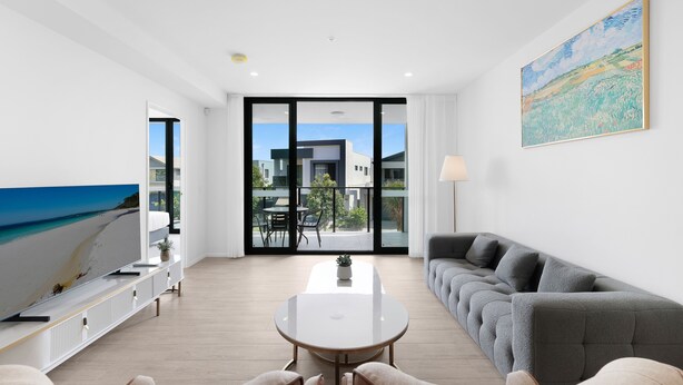 Uma sala de estar moderna com sofá, mesa de centro e TV. Há uma varanda com vista para edifícios e praia.