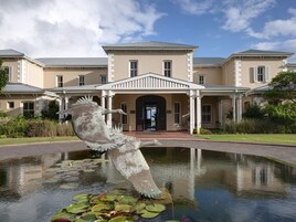 Um edifício grande com fachada simétrica, entrada central com pórtico e um lago com uma escultura em primeiro plano.