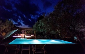 Piscina com espreguiçadeiras e área de deck à noite.