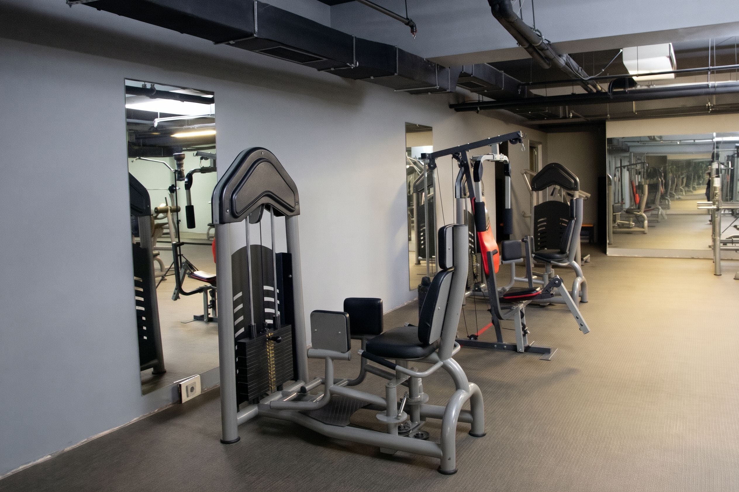 Academia com equipamentos de ginástica, espelhos e piso coberto de carpete.