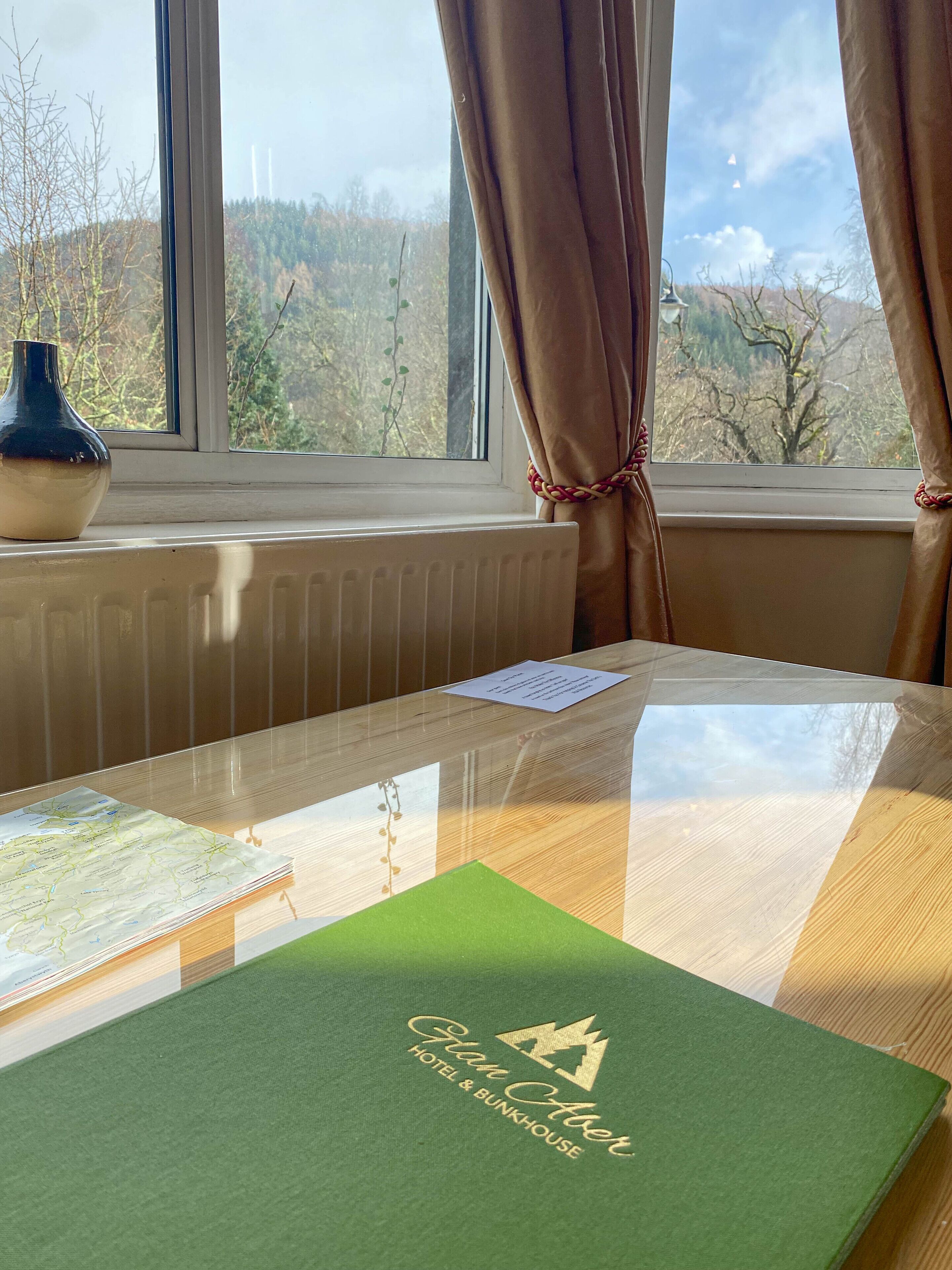 Quarto de hotel com vista para árvores e colinas pela janela, um radiador, uma mesa com um mapa e uma pasta verde, e um vaso no peitoril da janela.