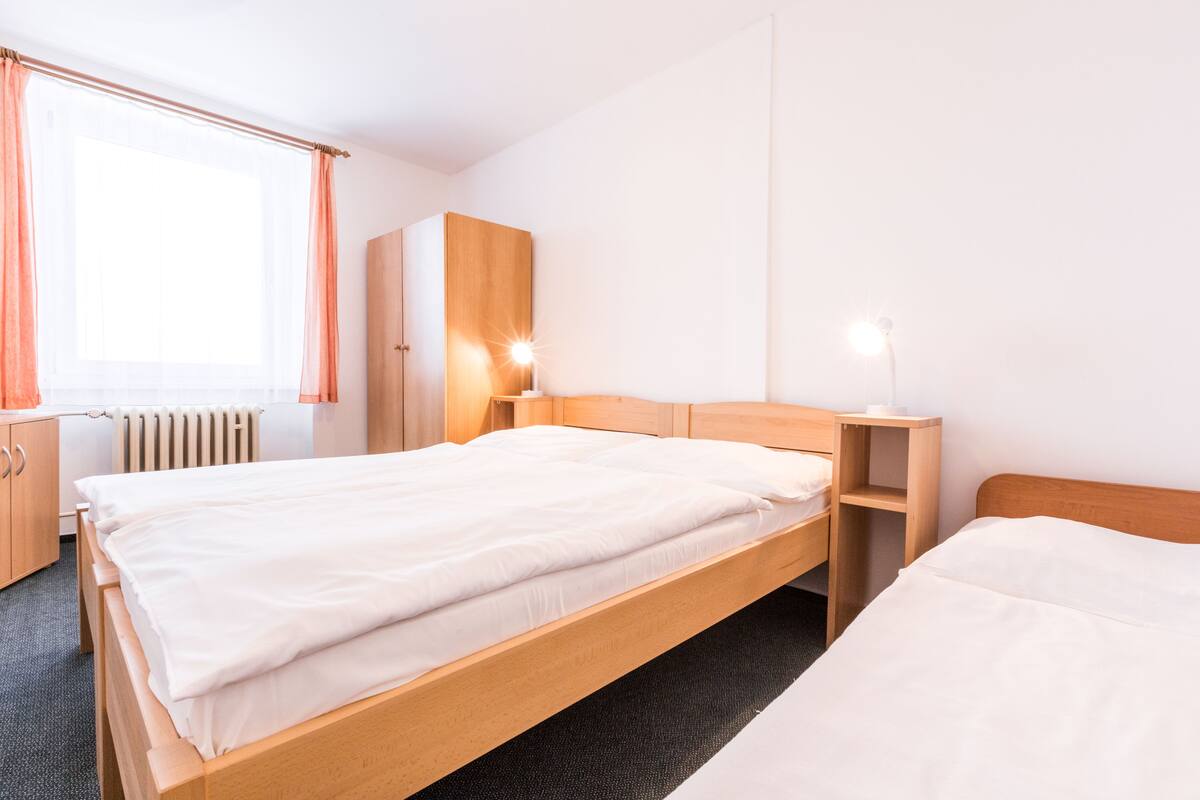 Quarto de hotel com uma cama de solteiro, guarda-roupa de madeira, mesa de cabeceira com abajur e janela com cortinas.