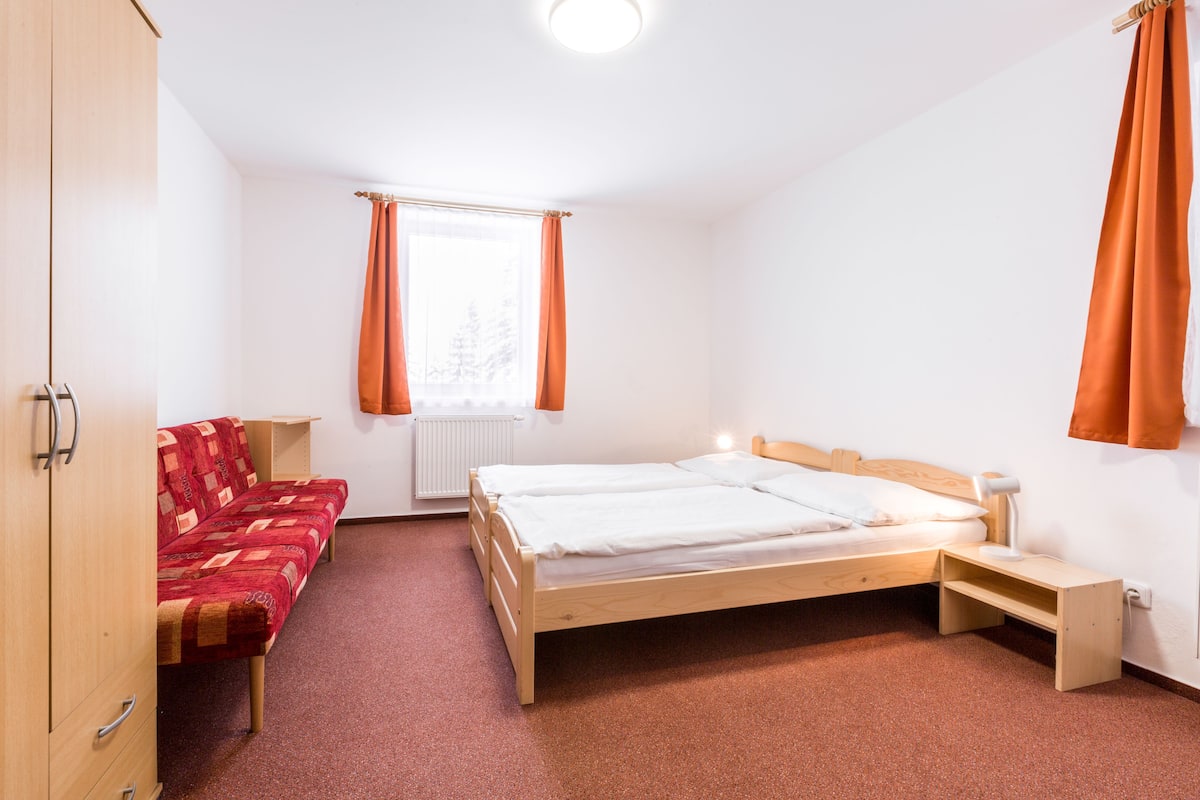 Quarto com duas camas de solteiro, um guarda-roupa de madeira, um sofá vermelho e uma janela com cortinas laranja.