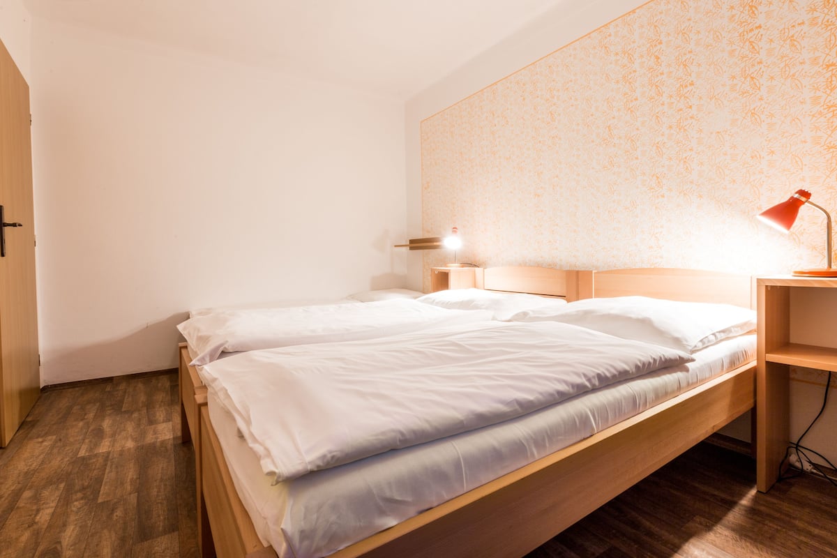Uma cama de solteiro com estrutura de madeira, roupa de cama branca e uma mesa de cabeceira com abajur vermelho.