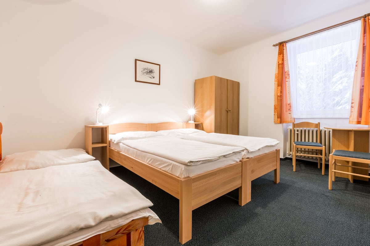 Quarto de hotel com duas camas de solteiro, um guarda-roupa de madeira, uma escrivaninha com cadeira e uma janela com cortinas.