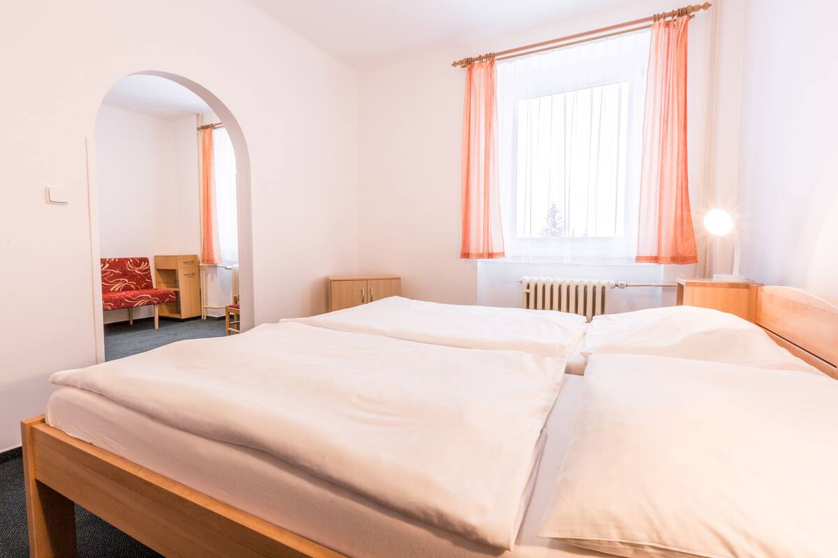 Quarto de hotel com duas camas, janela com cortinas, radiador e pequena mesa com abajur.