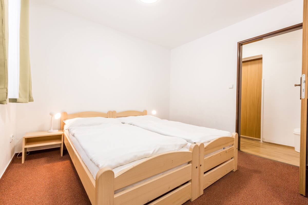 Quarto com cama de madeira, mesa de cabeceira com abajur e porta que dá para outro cômodo.