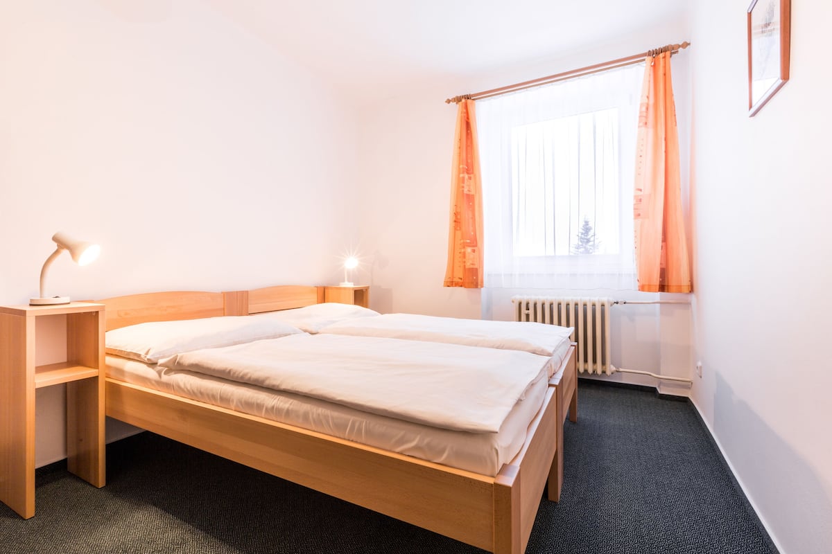 Uma cama bem arrumada com cabeceira de madeira, uma mesa de cabeceira com luminária, uma janela com cortinas laranja e um radiador.