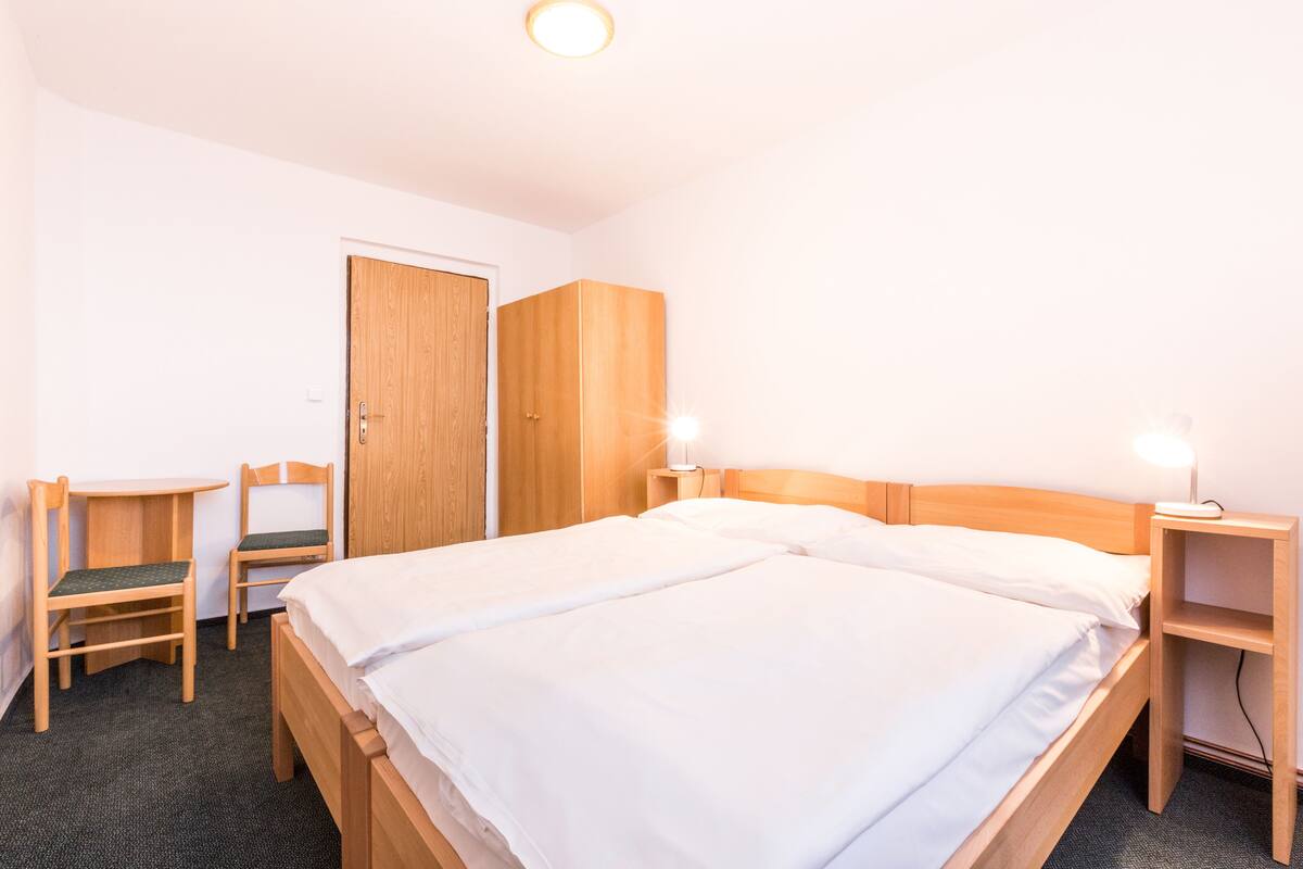 Quarto de hotel com cama de casal, guarda-roupa de madeira, secretária com cadeira e mesa de cabeceira com abajur.