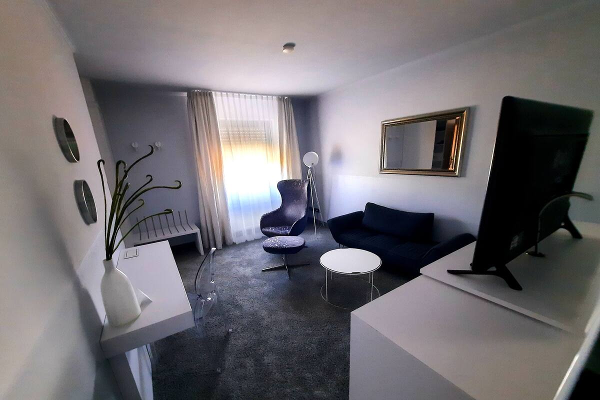 Um espaço de estar compacto com um sofá azul escuro, uma mesa de centro branca, uma mesa lateral branca com um vaso, um abajur e uma televisão em um suporte.