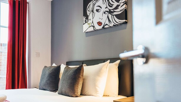 Quarto de hotel com cama, almofadas, mesa de cabeceira e uma obra de arte na parede.