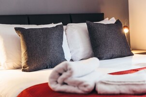 Cama bem arrumada com almofadas brancas e cinzentas, uma toalha dobrada e um cobertor vermelho.