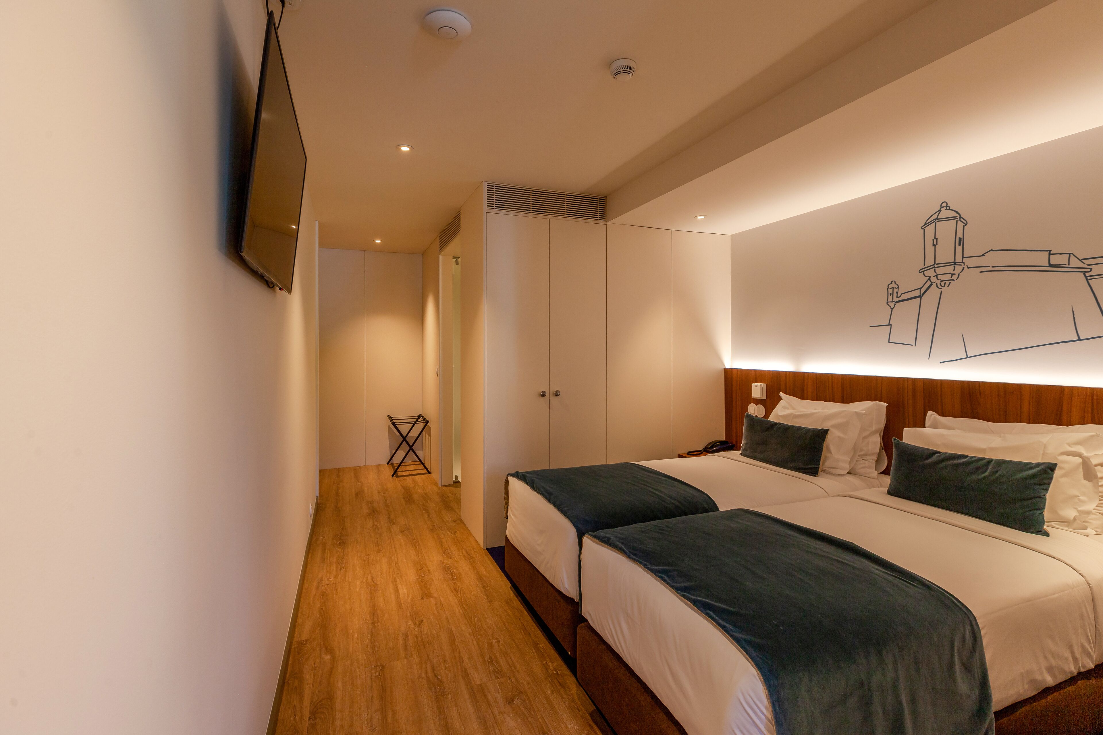 Quarto de hotel com duas camas, piso de madeira, televisão fixada na parede e ventilação no teto.