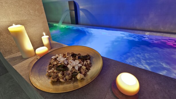 Espaço de spa com hidromassagem, velas e um prato de flores secas sobre superfície texturizada.