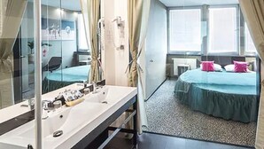 Quarto de hotel com um espelho grande, uma pia, uma cama com colcha verde e uma janela.