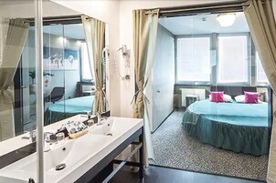 Quarto de hotel com um espelho grande, uma pia, uma cama com colcha verde e uma janela.