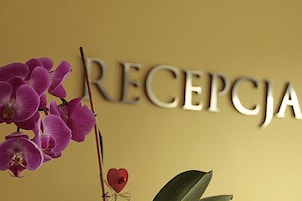 Placa de recepção com a palavra 'RECEPCJA' em letras grandes, acompanhada por uma orquídea roxa e um enfeite em formato de coração.