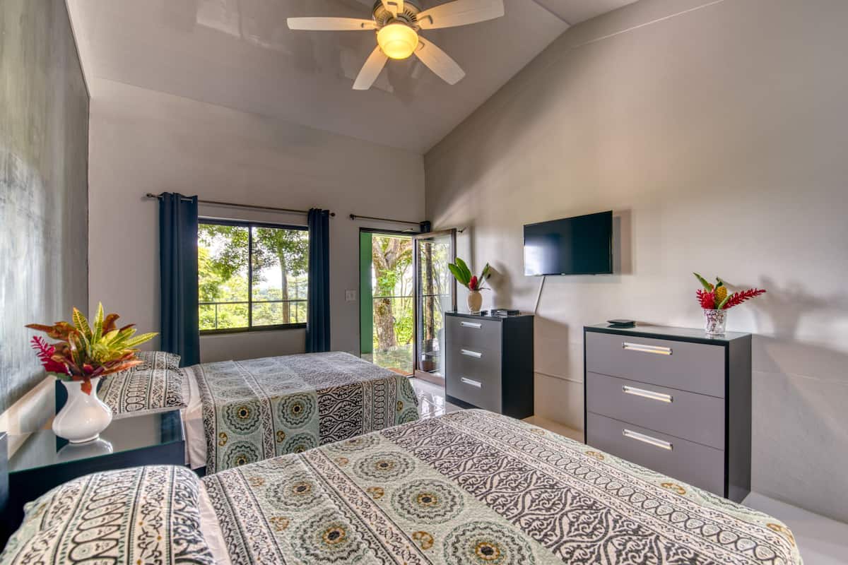 Quarto de hotel com duas camas, um ventilador de teto, televisão, cômoda e janela com cortinas.