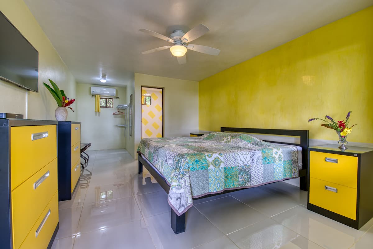 Quarto com cama, mesa de cabeceira, guarda-roupa, ventilador de teto e janela com unidade de ar condicionado.