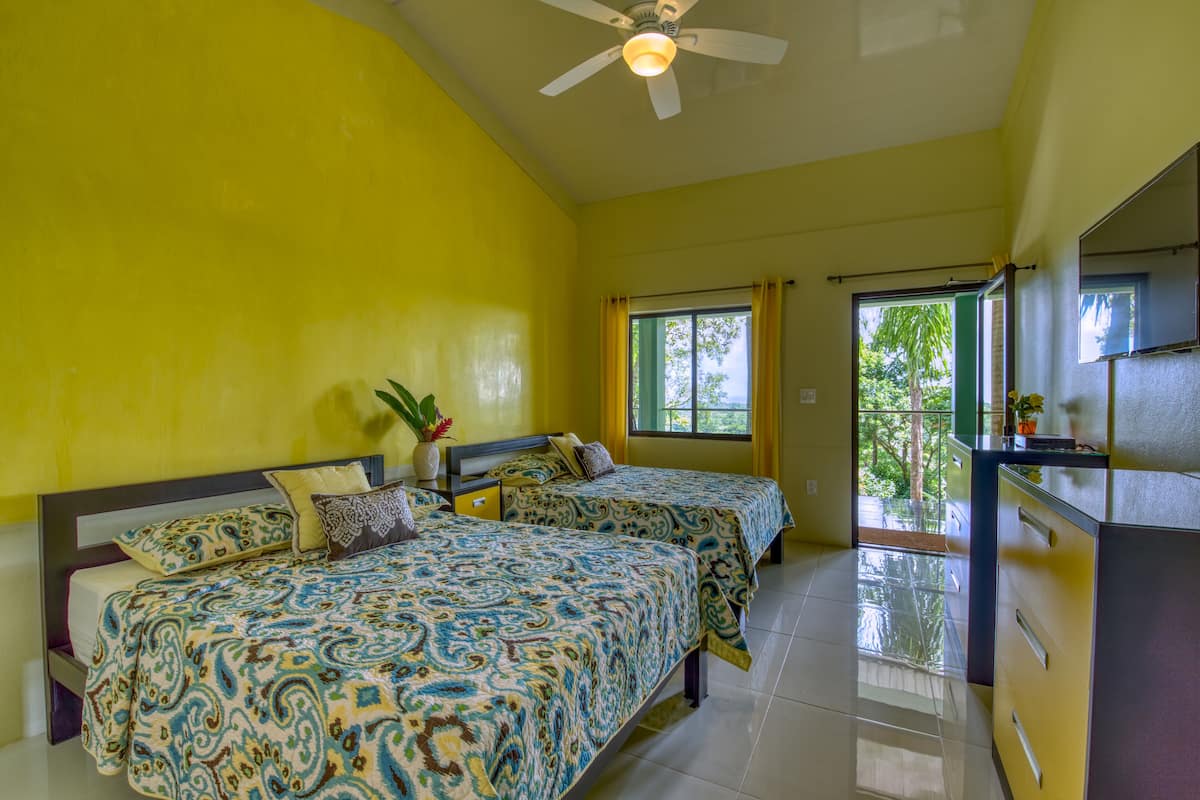 Quarto com duas camas, ventilador de teto e janela com cortinas.