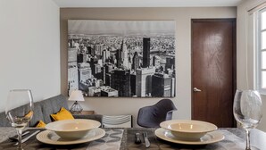 Uma área de refeições com vista para a cidade, uma mesa posta com pratos e uma taça de vinho, uma luminária e um sofá com almofadas.