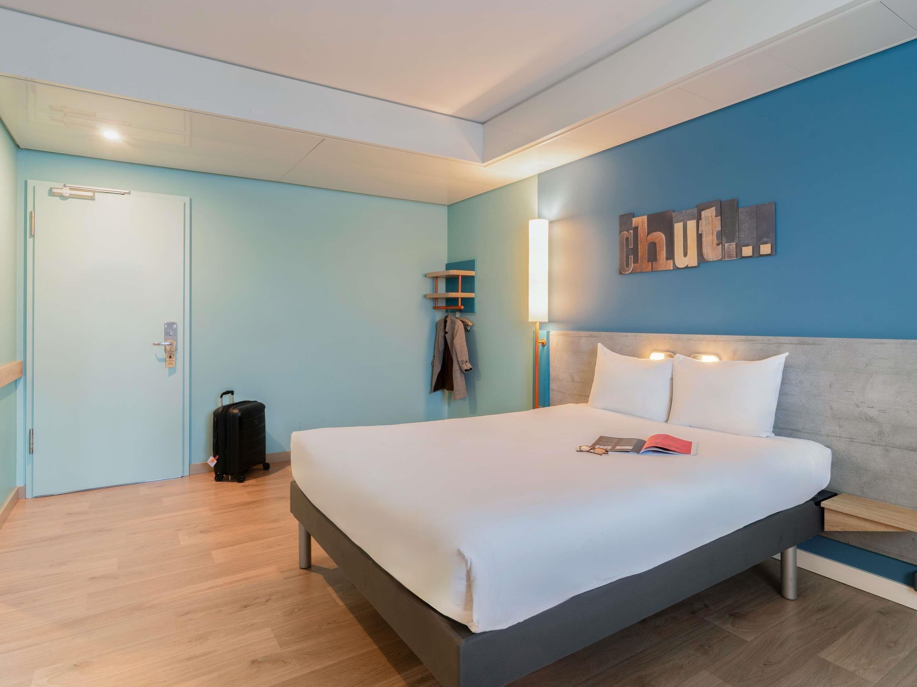 Quarto de hotel com cama, mala, cabideiro e parede com a palavra 'chute' parcialmente visível.