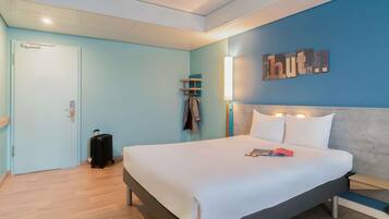 Quarto de hotel com cama, mala, cabideiro e parede com a palavra 'chute' parcialmente visível.