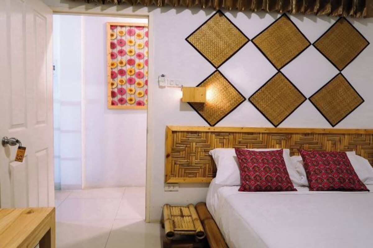 Quarto com cabeceira de madeira, cama com almofadas brancas e estampadas, luminária fixada na parede e painel decorativo com motivos florais.