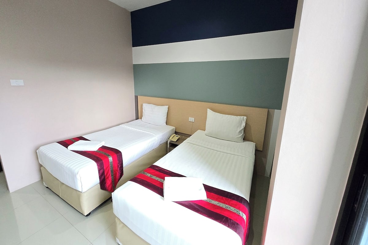 Quarto de hotel com duas camas de solteiro, cada uma com colchão branco e manta vermelha e bege. Há uma cabeceira de madeira, uma mesa de cabeceira com telefone e uma parede com padrão listrado.