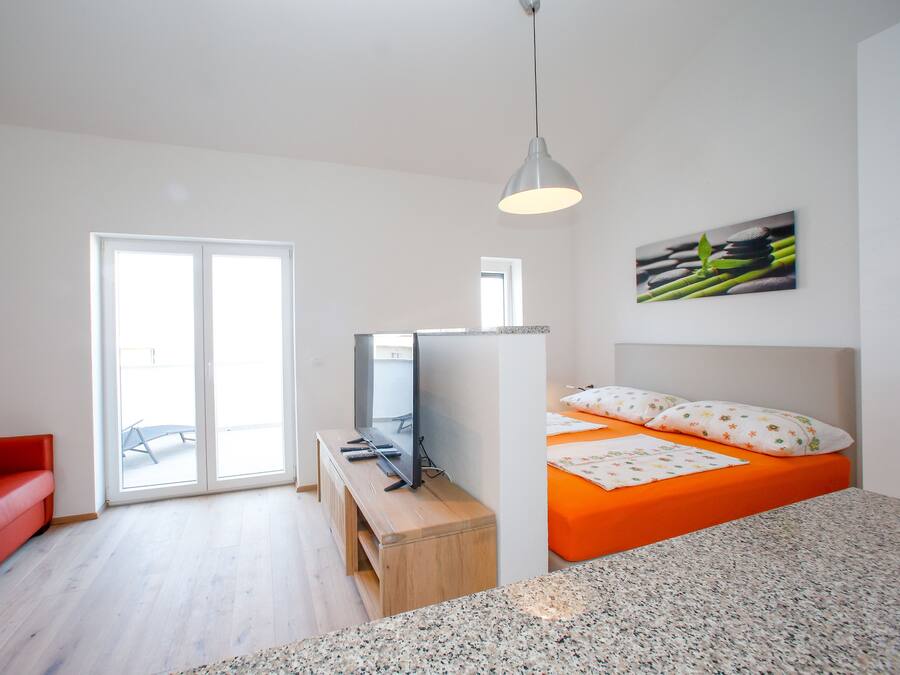 Quarto moderno com um sofá vermelho, uma cama com roupa de cama laranja, uma televisão sobre um suporte de madeira e um quadro na parede.