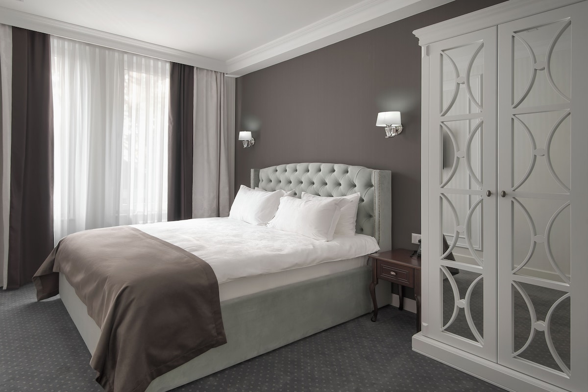 Uma cama bem arrumada com cabeceira cinzenta, uma mesinha de cabeceira com luminária e um guarda-roupa com painéis decorativos.