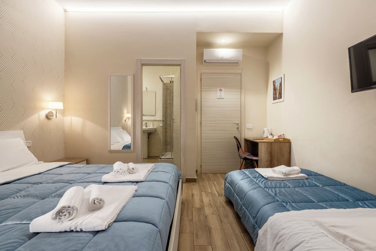 Quarto de hotel com duas camas, uma escrivaninha, uma cadeira, um espelho e área de chuveiro.